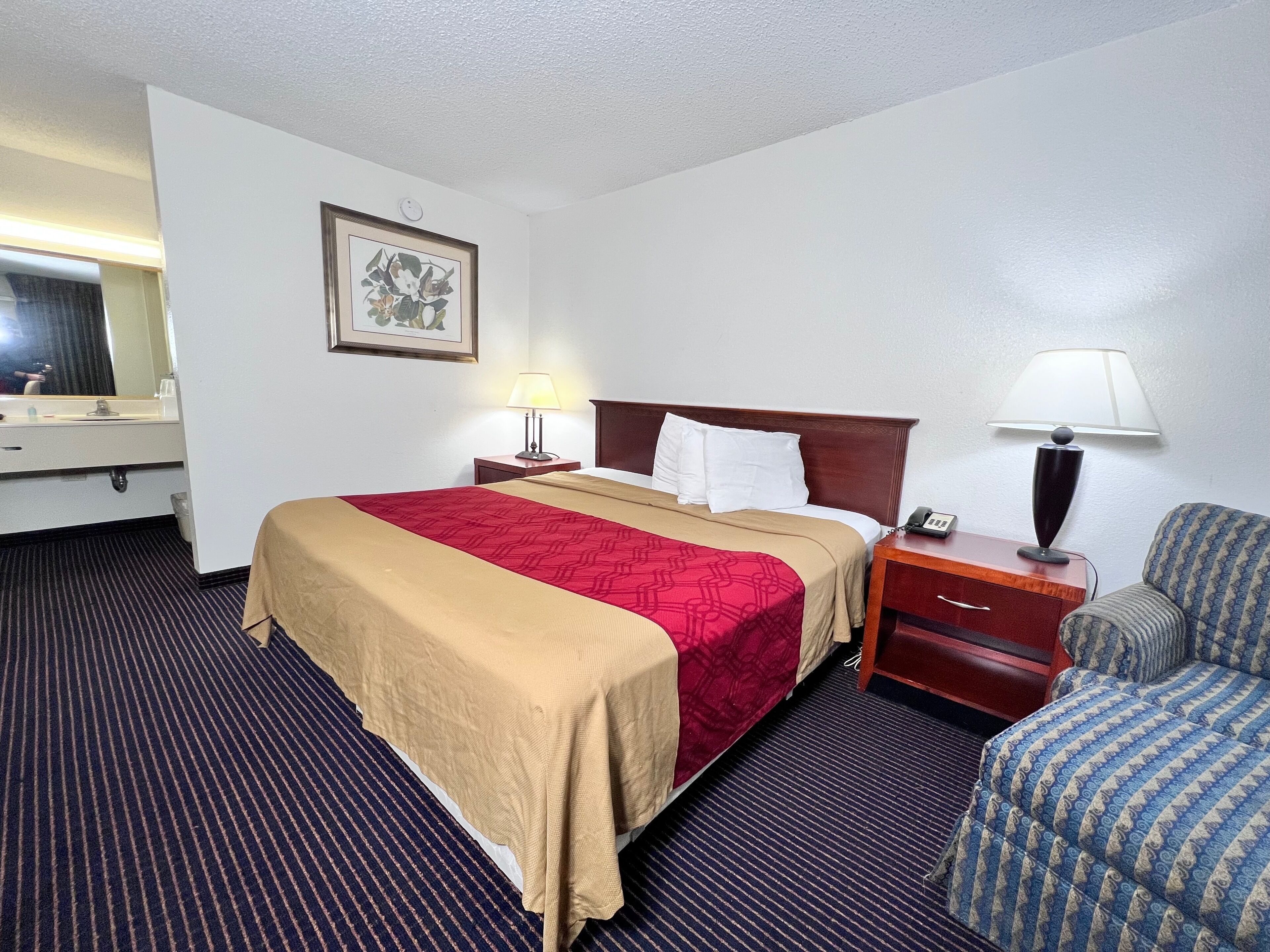 Quarto de hotel com cama, mesas de cabeceira, uma cadeira, espelho e um quadro na parede.