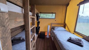 Quarto pequeno com uma cama de solteiro, um banquinho, uma prateleira com objetos e uma janela com vista.