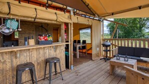 Uma área de bar ao ar livre com balcão de madeira, banquetas e um espaço de estar com sofá e mesa.