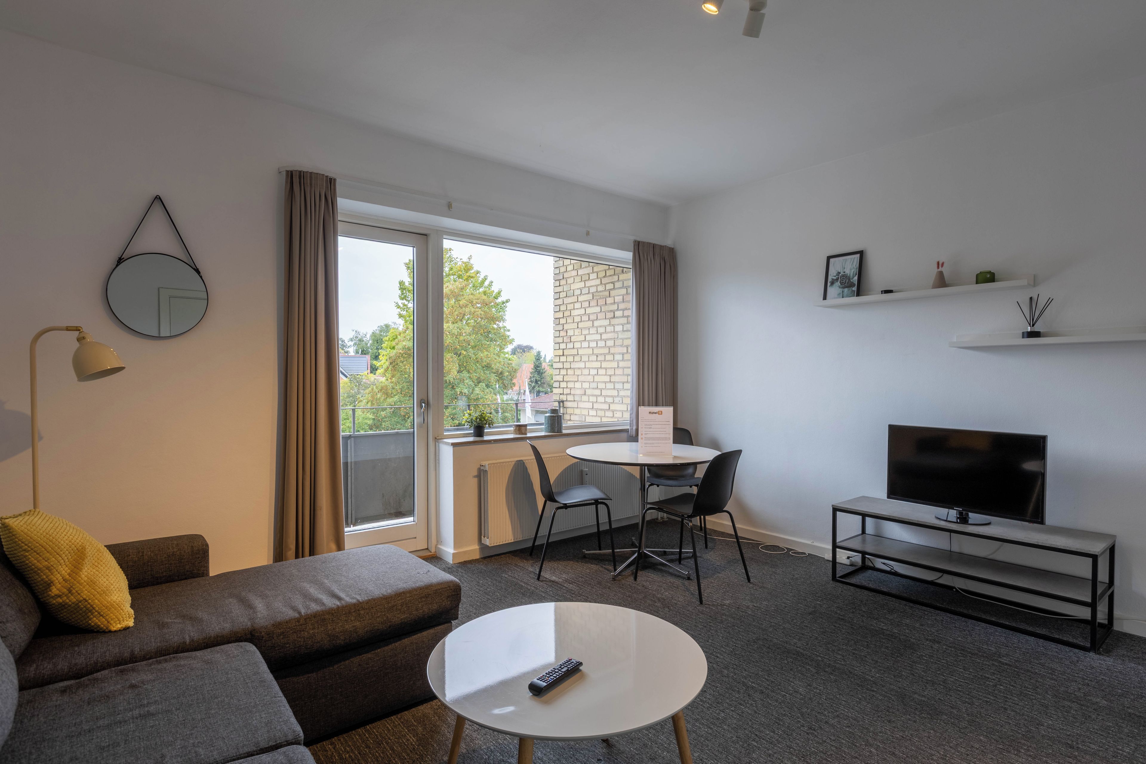 Sala de estar moderna com sofá, mesa de centro, televisão e varanda com vista para árvores e edifícios.