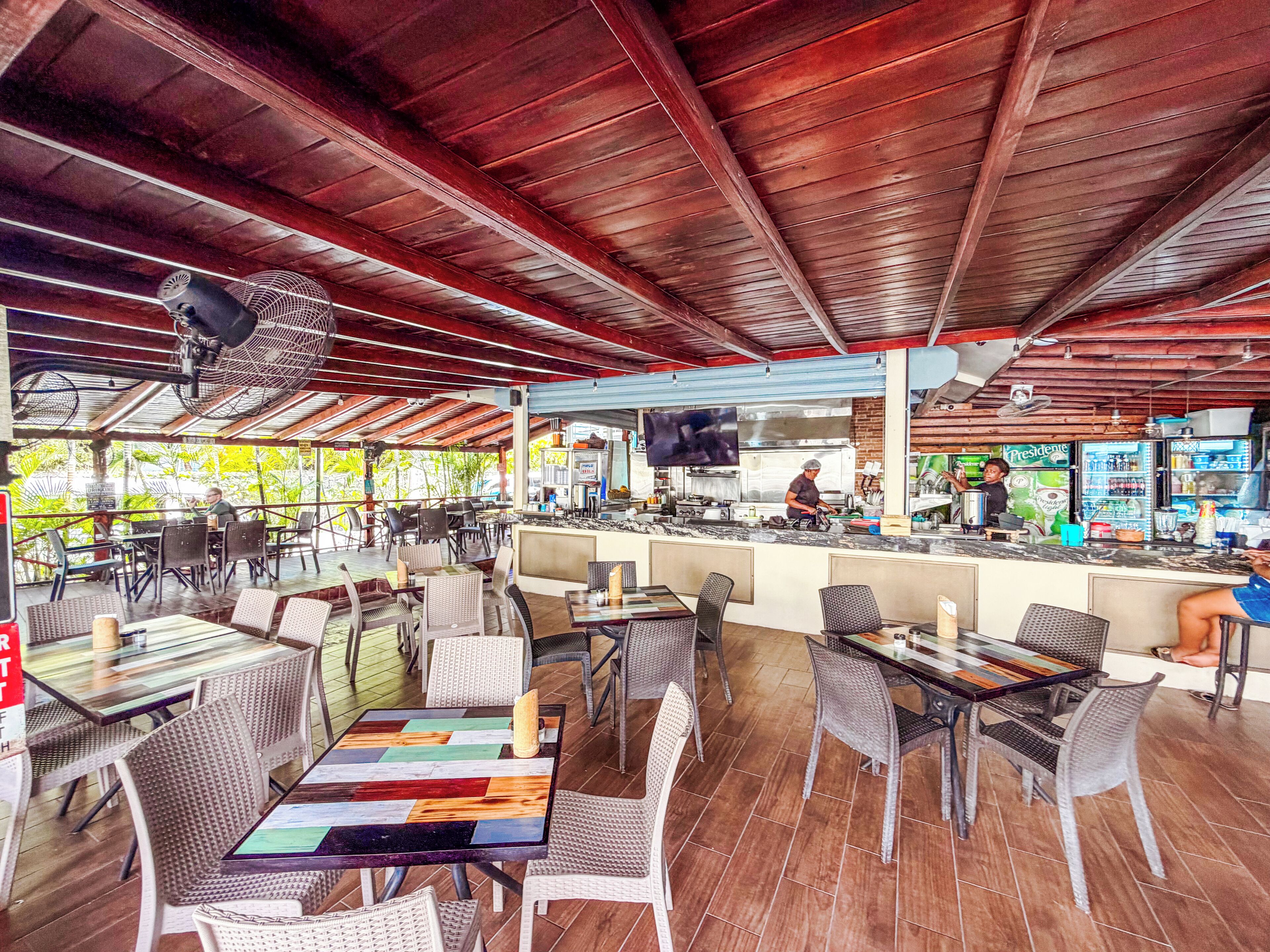 Restaurante ao ar livre com teto de madeira, mesas com jogos americanos coloridos e um espaço de bar com uma tela de televisão.