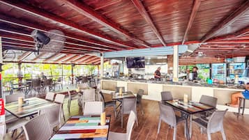 Restaurante ao ar livre com teto de madeira, mesas com jogos americanos coloridos e um espaço de bar com uma tela de televisão.