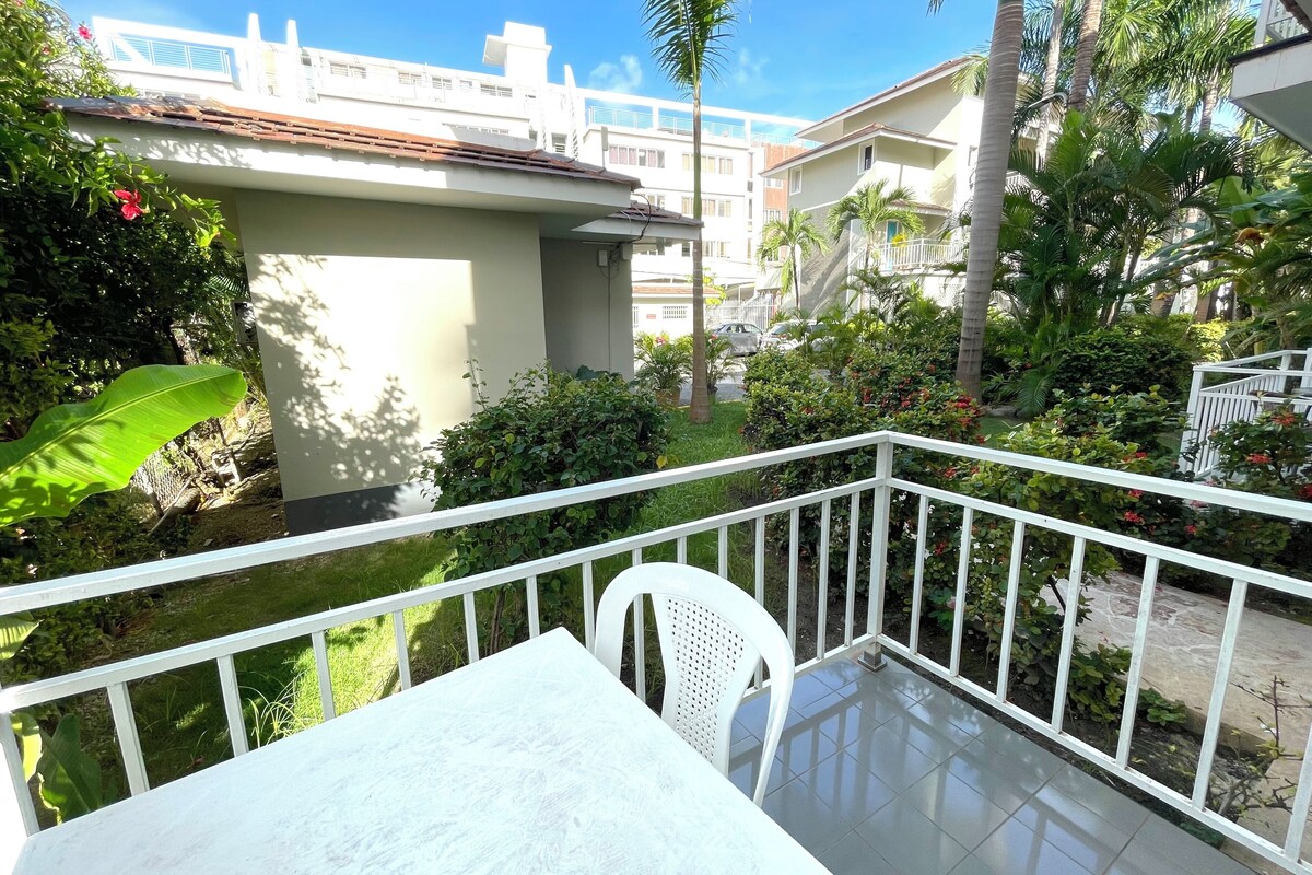 Varanda com mesa e cadeira brancas, cercada por vegetação exuberante e vista para edifícios vizinhos.