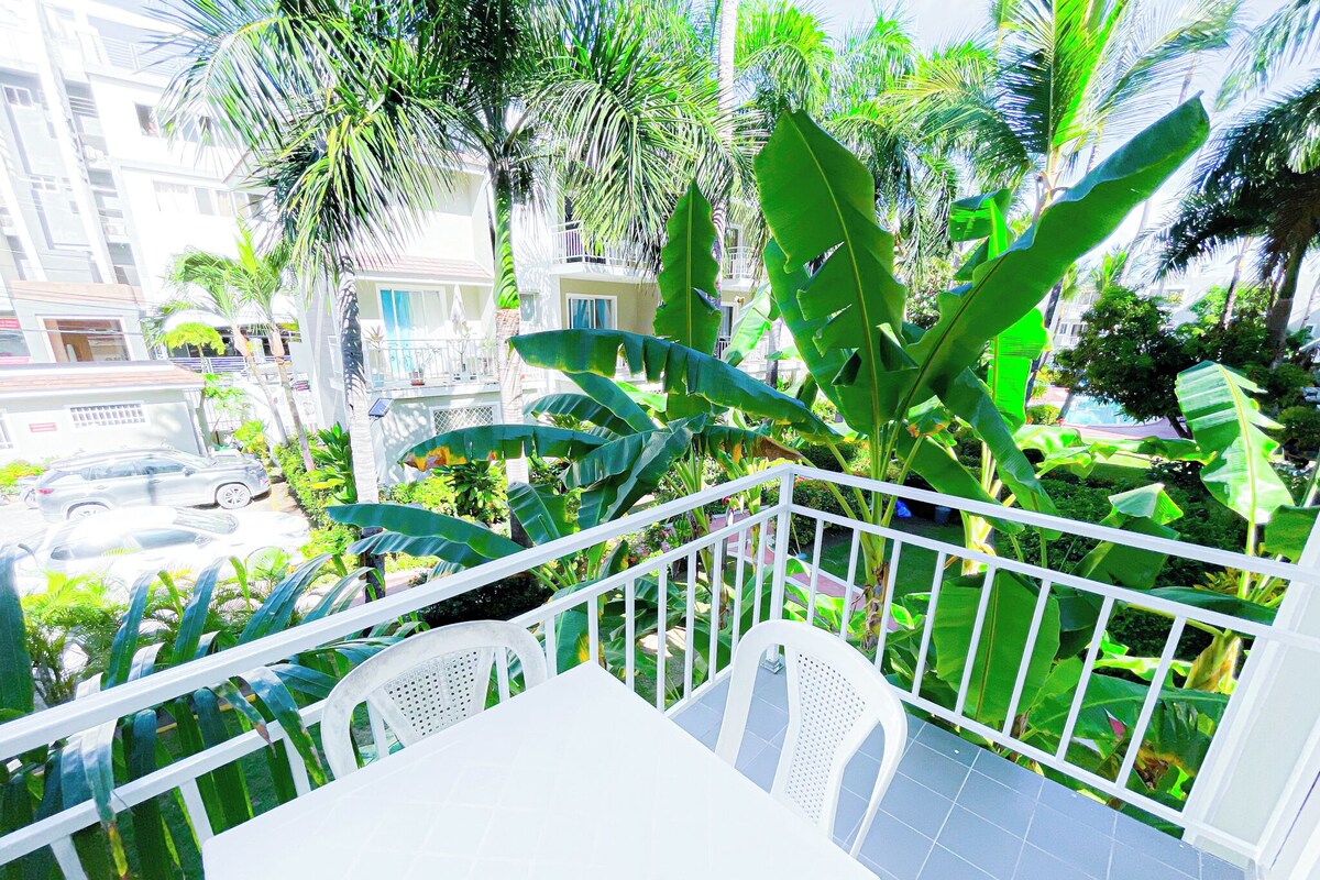 Varanda com cadeiras e mesa brancas, cercada por plantas verdes exuberantes e vista para uma área residencial.