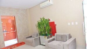 Um saguão de hotel com balcão de recepção, duas poltronas, uma planta em vaso e um aparelho de ar condicionado.