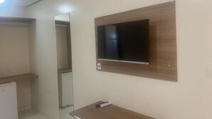 Quarto com televisão de tela plana presa a um painel de madeira, uma mesinha com controle remoto e um espelho na parede.