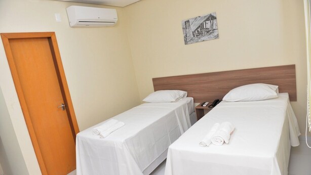 Quarto de hotel com duas camas de solteiro, uma porta de madeira, um aparelho de ar condicionado e uma pequena mesa com telefone.