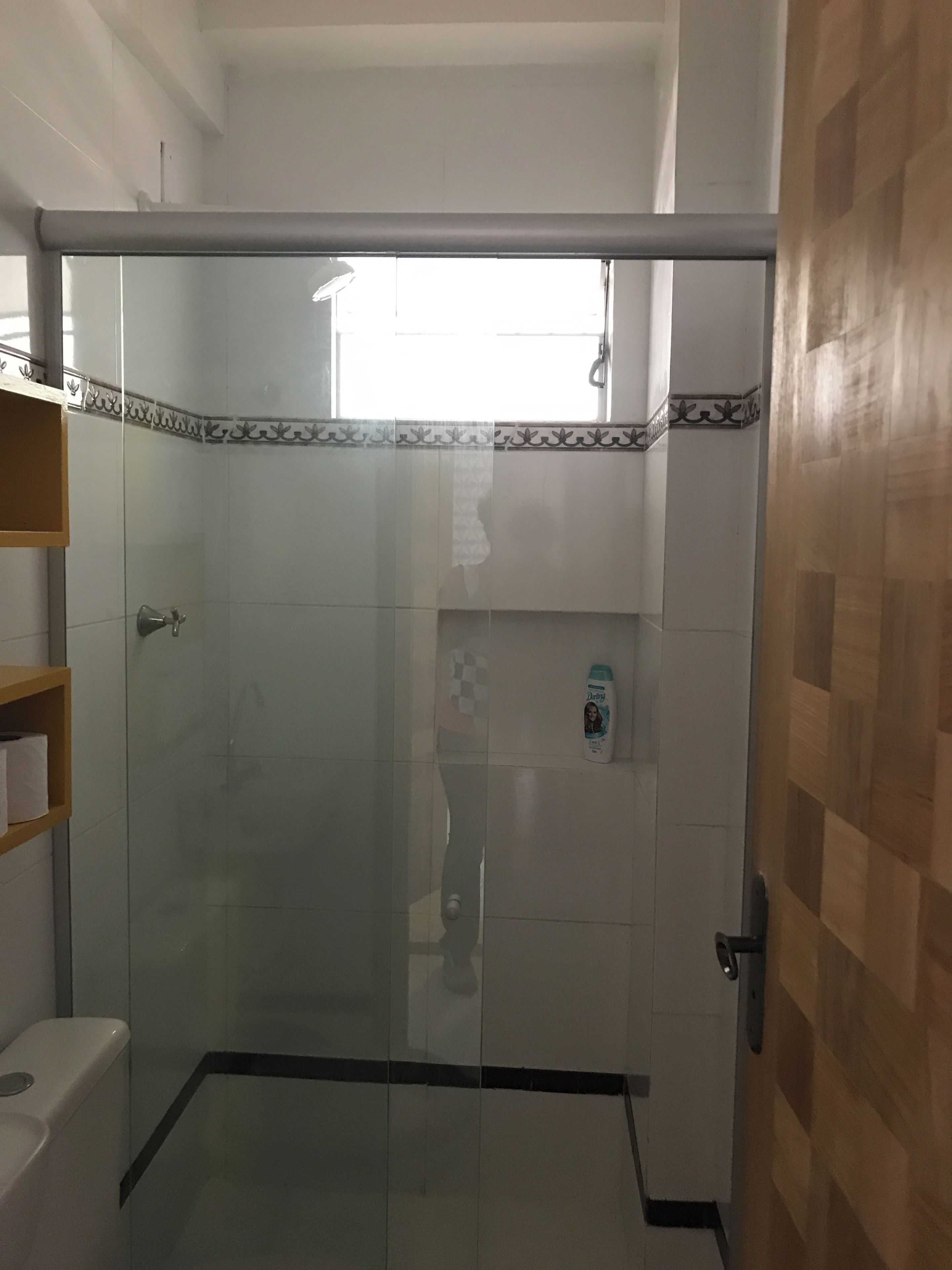 Área de chuveiro com paredes de vidro, piso de azulejo, prateleira de madeira e uma garrafa de shampoo.