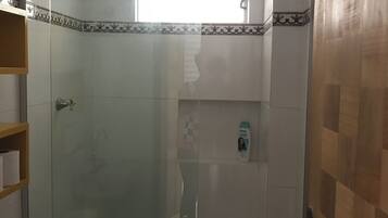 Área de chuveiro com paredes de vidro, piso de azulejo, prateleira de madeira e uma garrafa de shampoo.