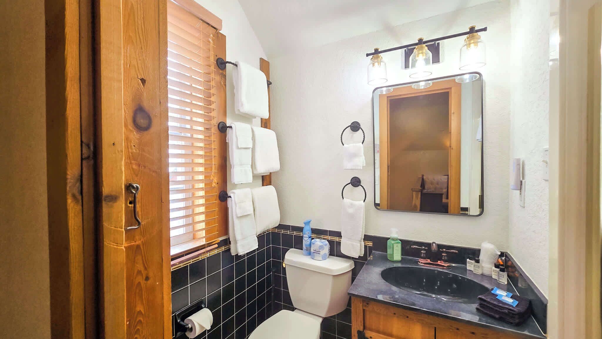 Uma casa de banho com porta de madeira, paredes azulejadas pretas, sanita branca, lavatório com artigos de higiene e um espelho com luzes.