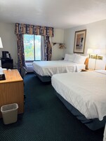 Quarto de hotel com duas camas, uma escrivaninha com cafeteira, um cesto de lixo e uma janela com cortinas.