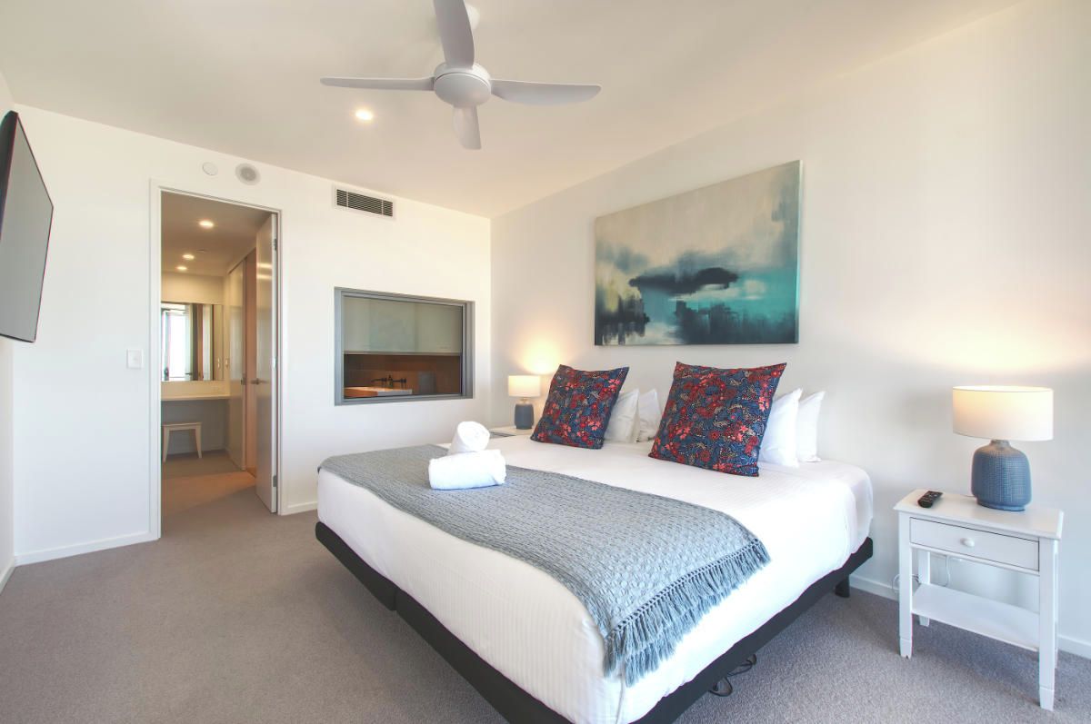 Quarto de hotel com cama, ventoinha no teto, um quadro, mesa de cabeceira e banheiro visível por uma porta aberta.