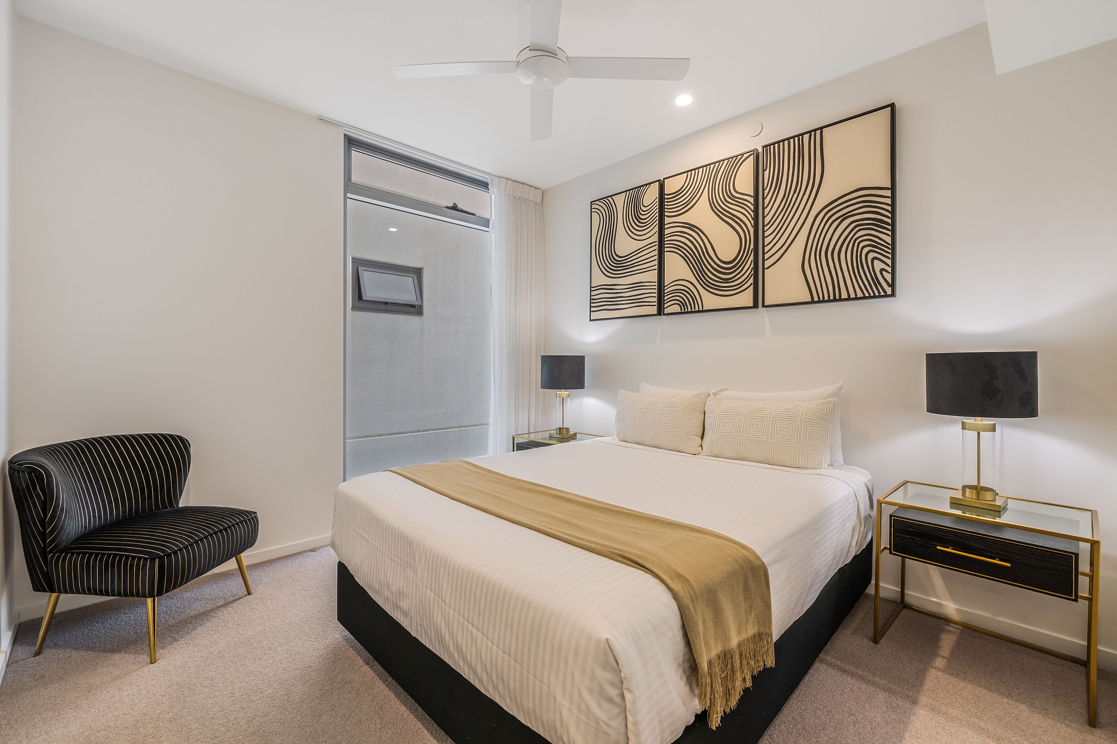 Quarto moderno com cama, mesas de cabeceira, uma cadeira e obra de arte na parede.