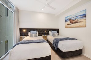 Quarto de hotel com duas camas, ventoinha no teto, pintura de uma cena de praia e janela com cortinas.