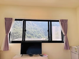 Quarto com uma janela grande, televisão de tela plana, uma mesa pequena com controle remoto e um refrigerador.