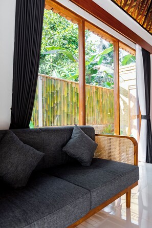 Uma sala de estar moderna com um sofá cinzento, uma poltrona de vime e uma janela ampla com vista para o verde.