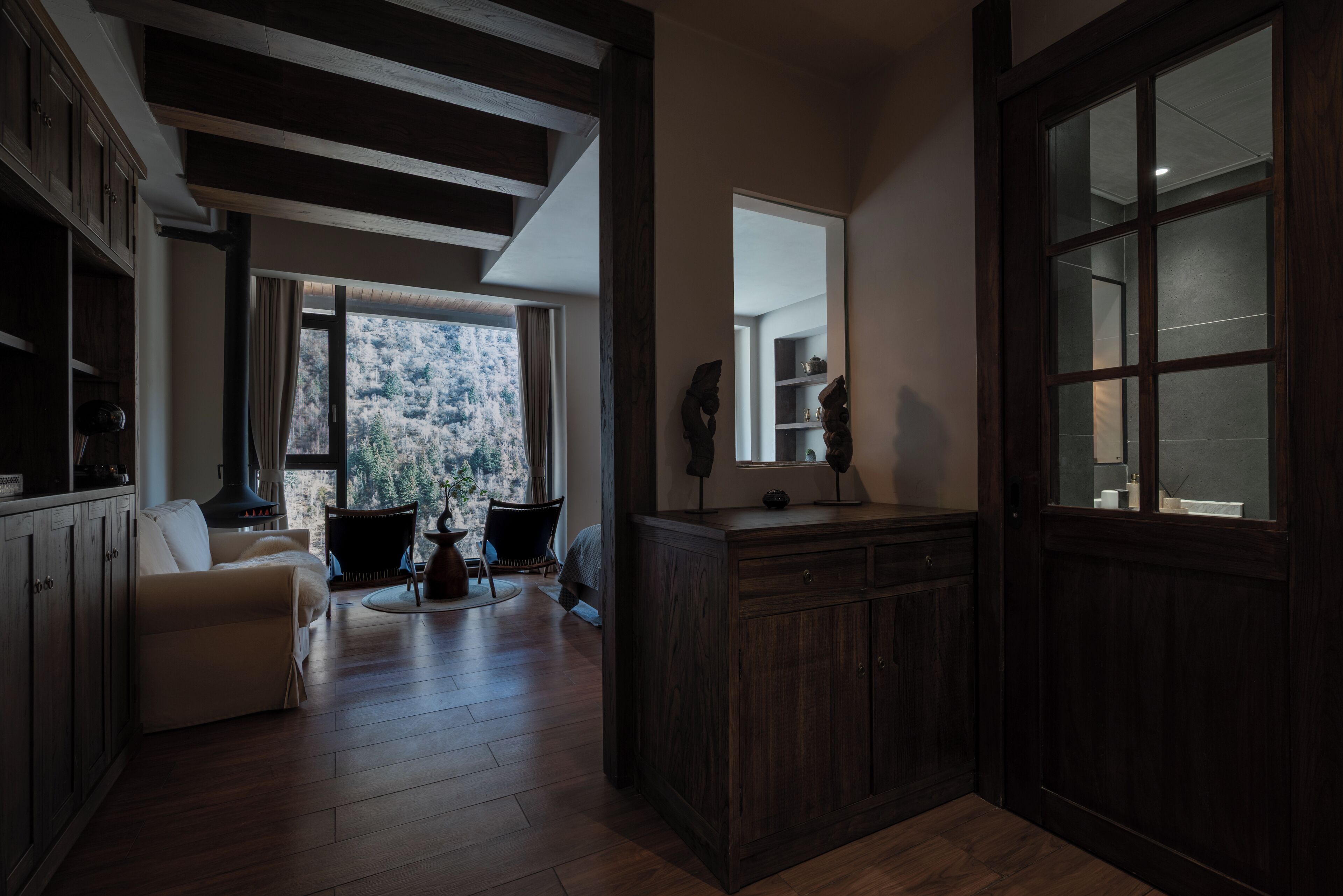 Quarto com piso de madeira, sofá branco, armário de madeira e uma janela grande com vista para uma área arborizada.