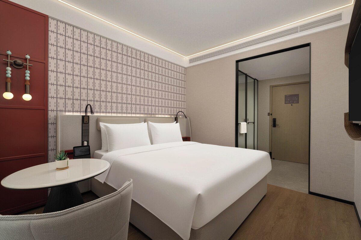Quarto de hotel com uma cama grande, mesa de cabeceira, uma cadeira, uma mesinha e televisão.
