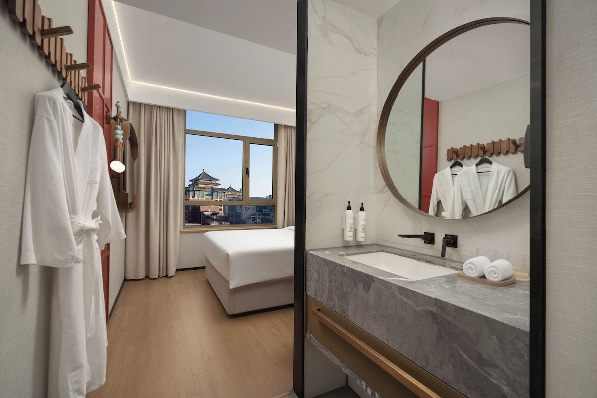 Quarto de hotel com cama, banheiro com pia e espelho, e vista para edifícios tradicionais pela janela.