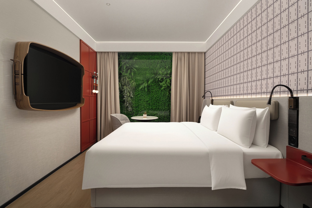Quarto de hotel com cama, televisão, secretária e vista para área verde pela janela.