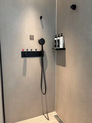 Área de chuveiro com chuveiro manual, painel de controle com vários botões e prateleira com artigos de higiene pessoal.