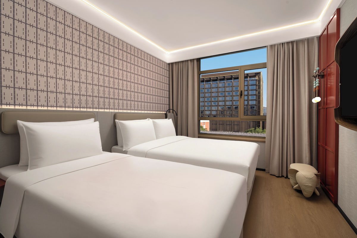 Quarto de hotel com duas camas, uma janela grande com vista para edifícios e uma televisão na parede.