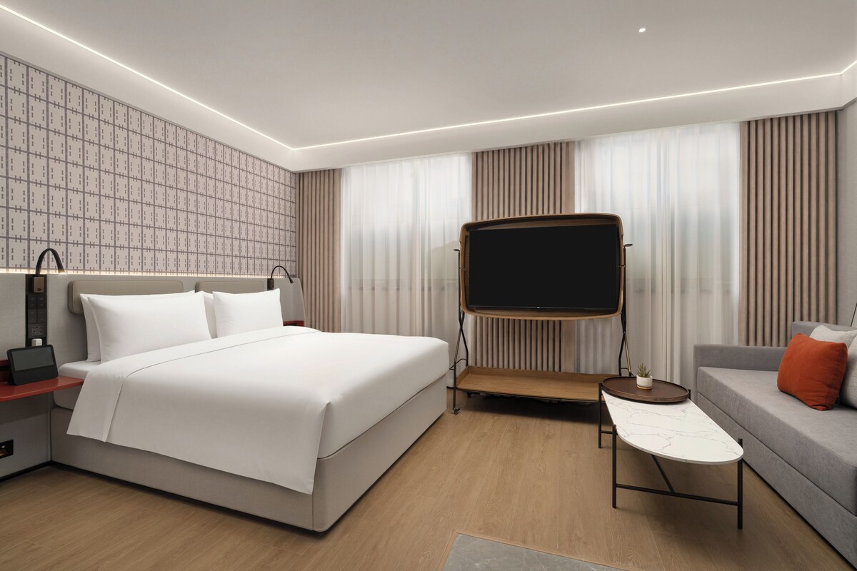 Quarto de hotel moderno com uma cama grande, uma mesa de cabeceira com abajur, uma TV em um suporte, um sofá e uma pequena mesa.