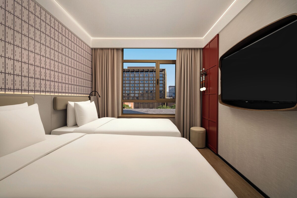 Quarto de hotel com duas camas, uma janela grande, uma TV de tela plana e uma TV fixada na parede.