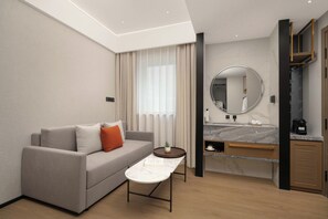Quarto de hotel moderno com sofá cinzento, uma pequena mesa, um espelho redondo e casa de banho com bancada de mármore.