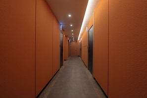 Um corredor comprido e estreito, com paredes laranja e chão cinzento, que leva a uma porta no final.