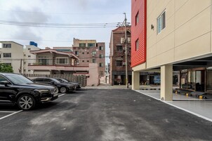 Um estacionamento com vários carros, um edifício com fachada vermelha e outros edifícios residenciais ao fundo.