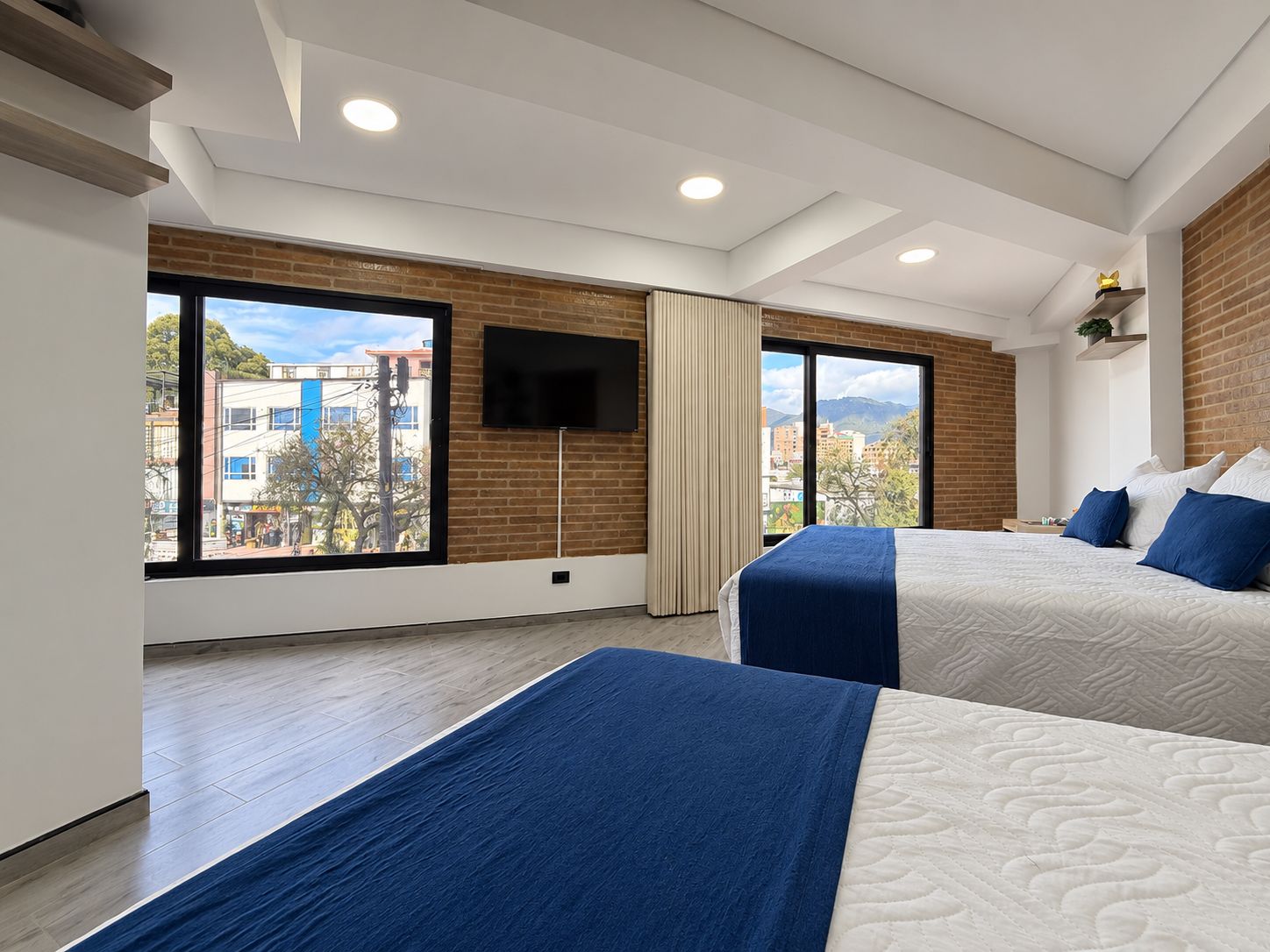 Quarto de hotel com duas camas, televisão na parede e grandes janelas com vista para a cidade.