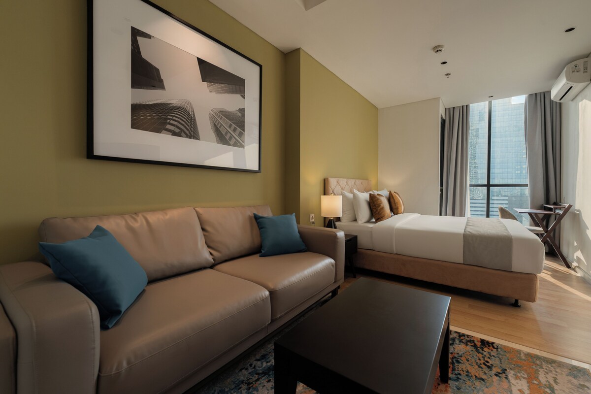 Quarto de hotel com cama, sofá, escrivaninha e uma janela grande.