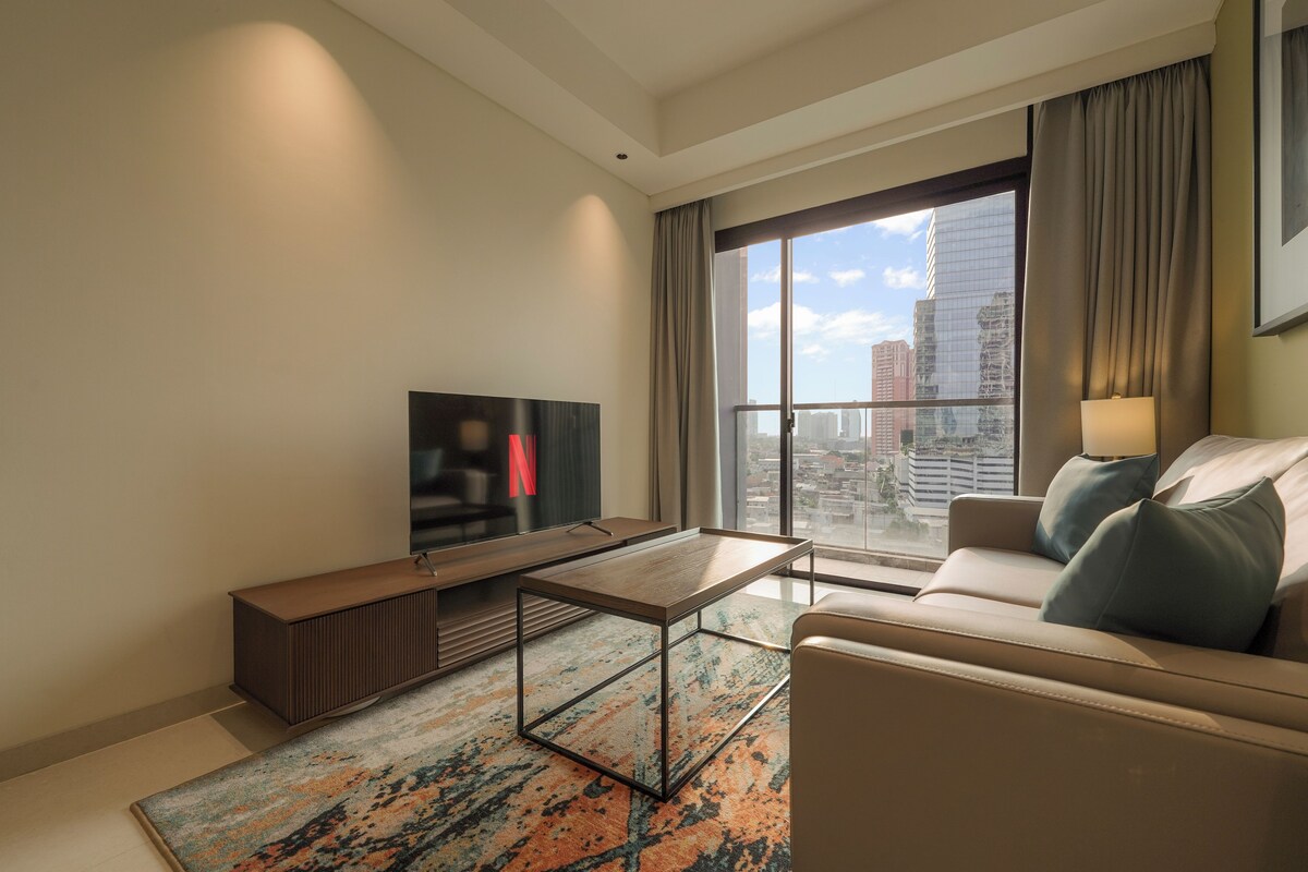 Uma sala de estar moderna com televisão de tela plana, sofá, mesa de centro e uma grande janela com vista para a cidade.