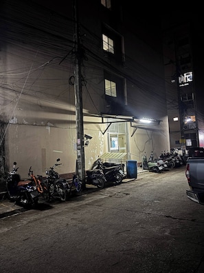Uma área de estacionamento à noite, com motocicletas e scooters estacionados em frente a um edifício com sinais de desgaste.