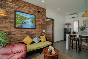 Uma sala de estar com parede de tijolos, um sofá com almofadas, uma mesa de centro com objetos decorativos e um quadro de uma paisagem fluvial.