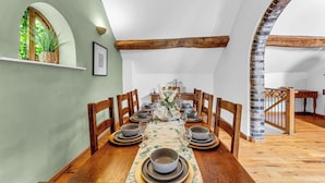 Uma área de refeições com uma mesa de madeira posta para seis pessoas, rodeada de cadeiras, com vista para um jardim pela janela.
