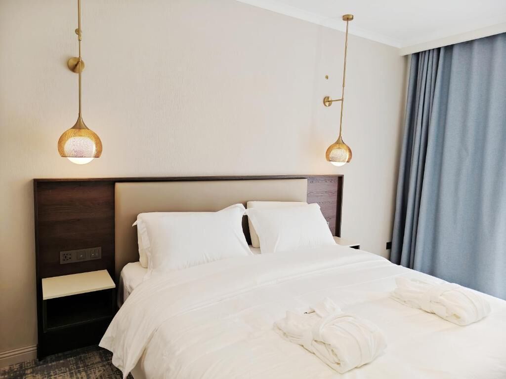 Quarto de hotel com cama, duas luminárias pendentes, uma mesinha de cabeceira e janela com cortinas.