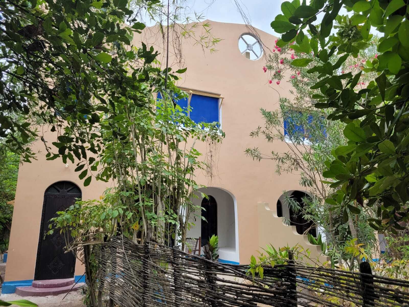 Um edifício com várias arcadas e uma janela circular, cercado por vegetaç ão exuberante e um cercado de palha.
