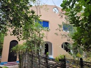 Um edifício com várias arcadas e uma janela circular, cercado por vegetação exuberante e um cercado de palha.