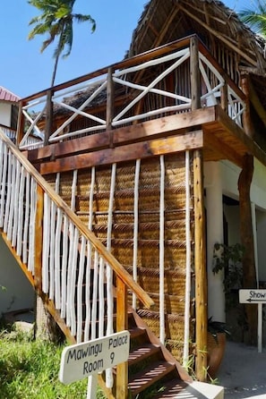 Uma escada de madeira leva a um quarto com telhado de palha e uma placa que indica 'Mawingu Palace Room 9'.