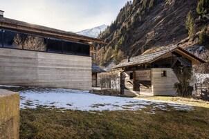 Uma cabana rústica com telhado inclinado, rodeada por terreno coberto de neve e uma paisagem montanhosa ao fundo.