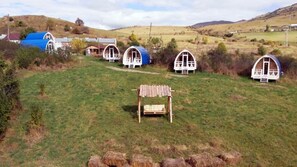 Um cenário rural com várias cabanas pequenas em formato de cúpula, com grades brancas, um banco de madeira e fardos de feno em um campo gramado.