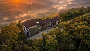 Um edifício de dois andares com telhado plano, cercado por uma floresta densa e vista para colinas distantes.