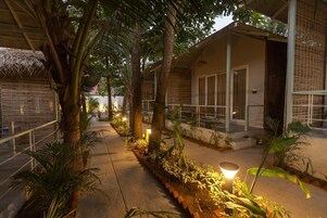 Uma trilha bordeada por árvores e plantas que leva a um edifício com telhado de palha e varanda.
