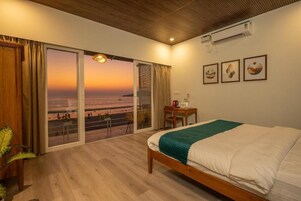 Quarto de hotel com cama, escrivaninha, cadeira, vista para o mar e ventoinha no teto.
