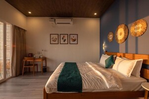 Quarto de hotel com cama, escrivaninha e cadeira. Há unidades de ar condicionado fixadas na parede e arte decorativa.