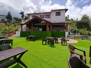 Uma área externa com mesas e cadeiras, um edifício com telhado vermelho e um gramado verde.