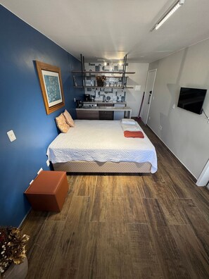 Quarto com cama, pequeno espaço de cozinha, televisão e piso de madeira.