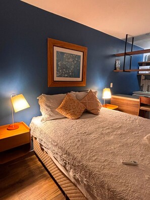 Quarto com cama, duas luminárias de cabeceira, um quadro na parede e uma mesinha de cabeceira de madeira.